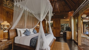 Uma cama de madeira com dossel, roupa de cama branca e cortina, uma mesinha de cabeceira com abajur e vista para o exterior por uma porta de vidro.