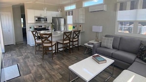 Uma sala de estar moderna com um sofá cinzento, uma mesa de centro e um espaço de jantar com cadeiras de madeira e uma mesa branca.