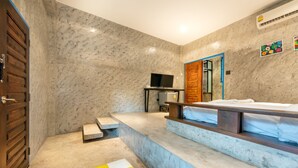 Quarto de hotel com parede de mármore, cama com roupa de cama branca, televisão sobre suporte e pequena mesa com cadeira.