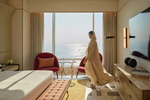 Quarto de hotel com uma cama grande, uma poltrona vermelha, uma mesinha com um copo e vista para o oceano.