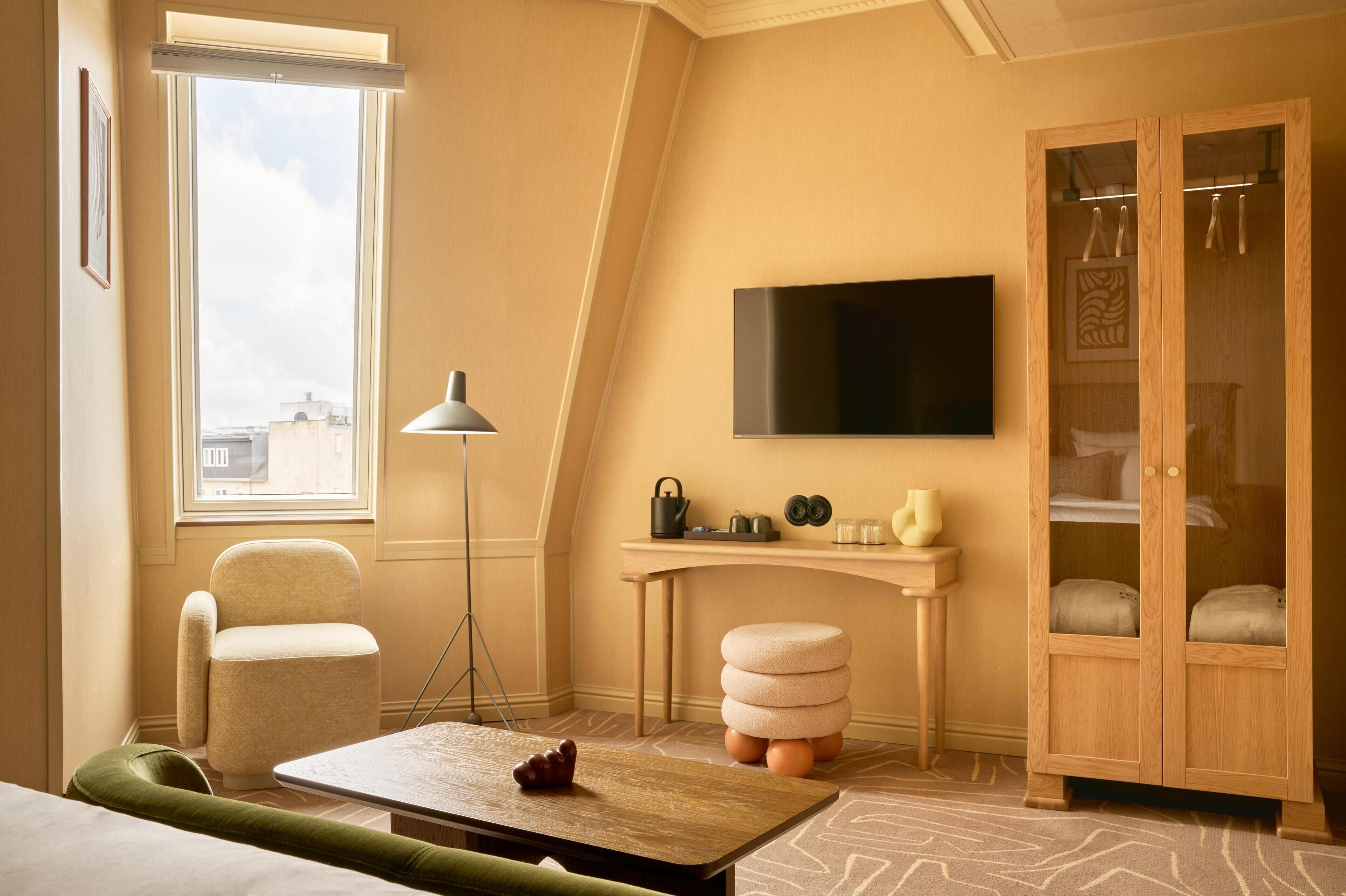 Um quarto acolhedor com uma janela grande, televisão de tela plana, guarda-roupa de madeira, uma mesa pequena e um sofá verde.