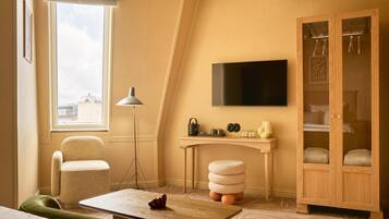 Um quarto acolhedor com uma janela grande, televisão de tela plana, guarda-roupa de madeira, uma mesa pequena e um sofá verde.