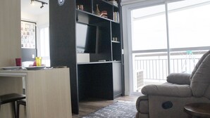 Uma sala de estar com sofá, área de jantar com mesa e cadeiras, estante de livros e uma janela grande com vista para o exterior.