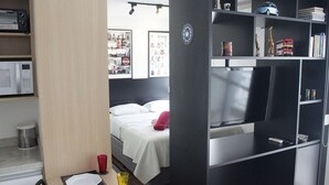 Quarto de hotel com uma cama, televisão, micro-ondas e uma estante com diversos objetos.