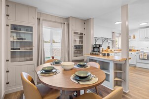 Uma sala de jantar com uma mesa redonda, cadeiras e uma cozinha ao fundo.