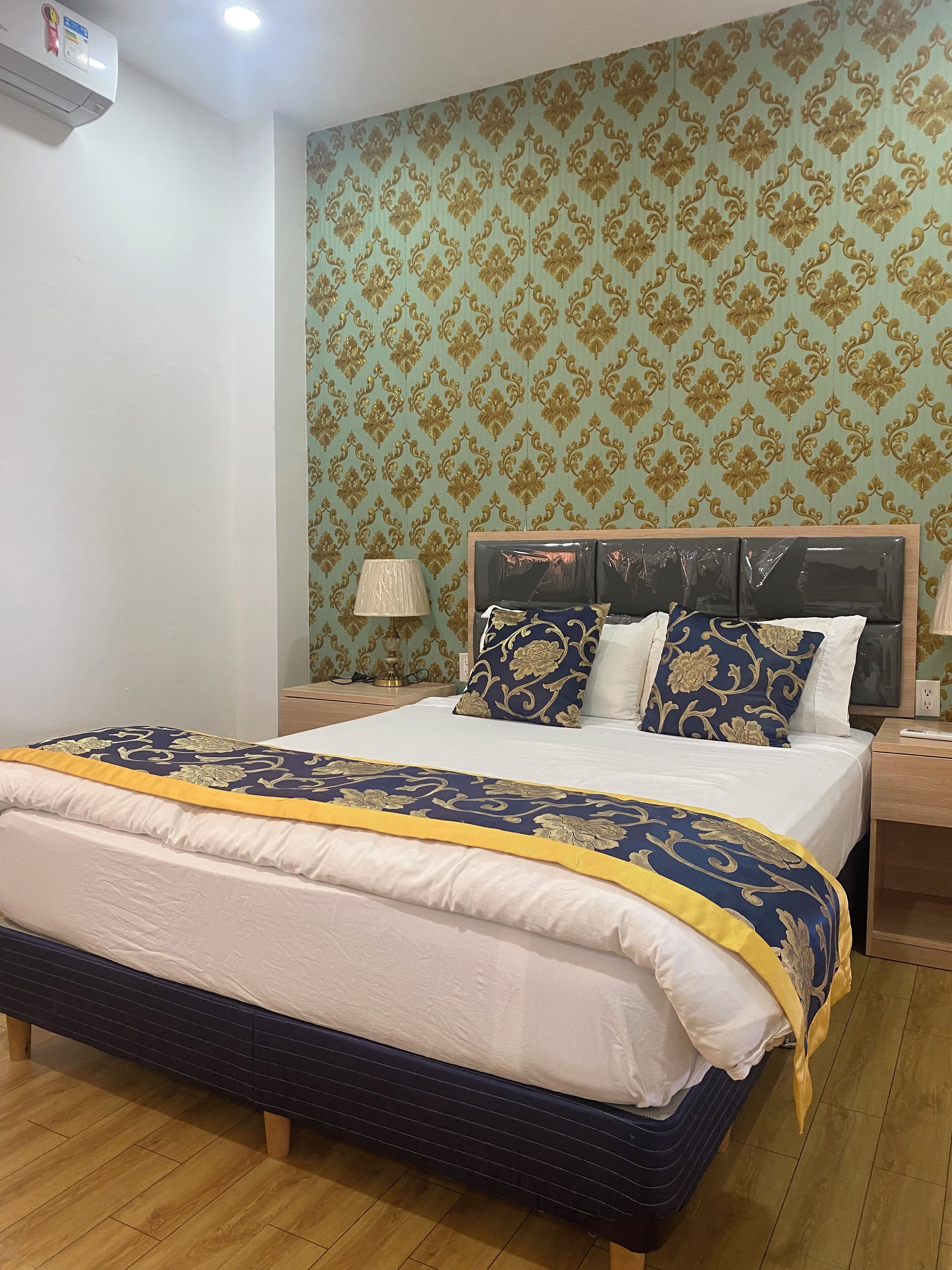 Uma cama bem arrumada com colcha azul e amarela, cabeceira de madeira e mesa de cabeceira com abajur.