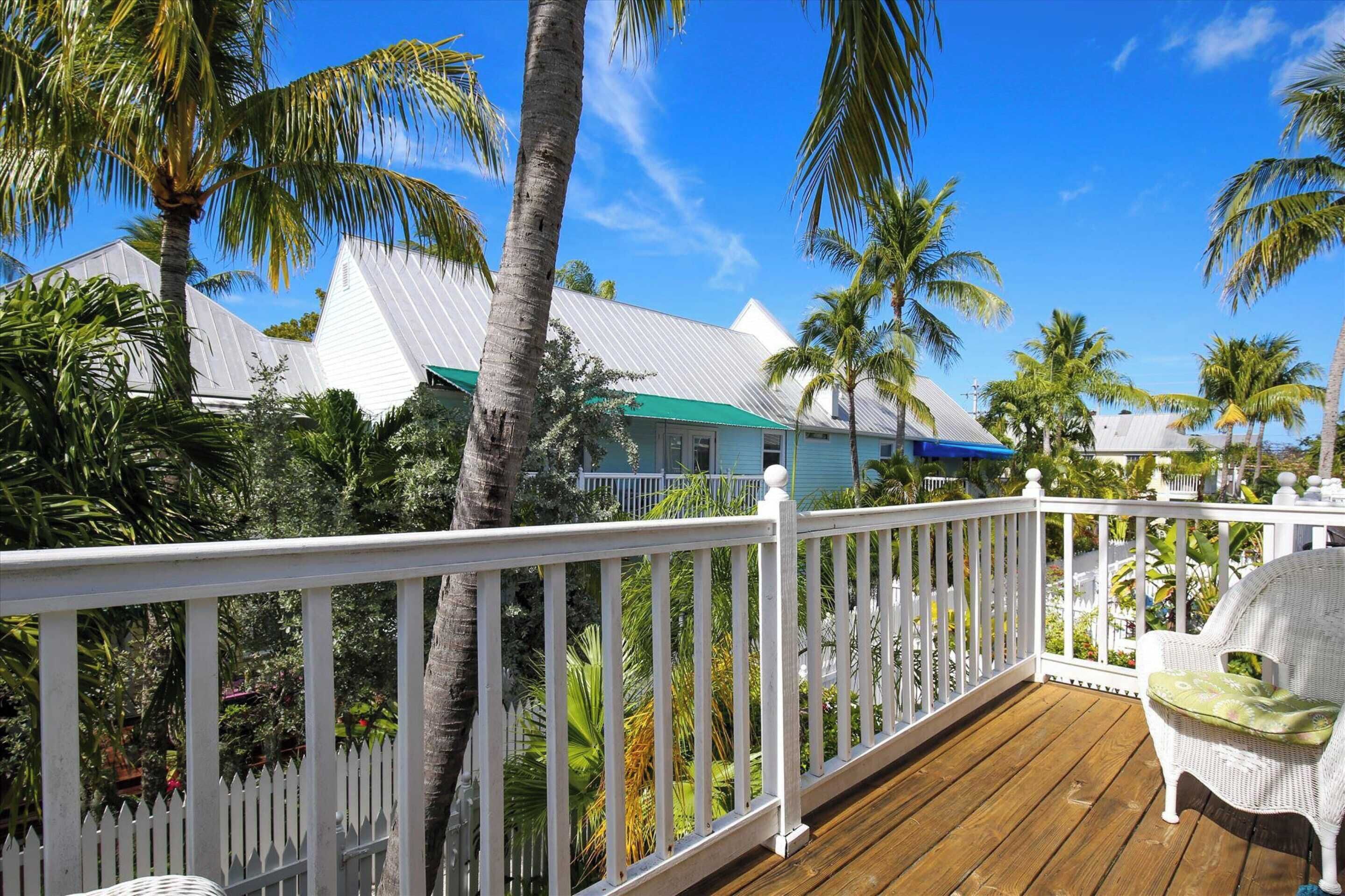 Um deck de madeira com guarda-corpo branco, uma cadeira de vime e vista para uma área de resort com palmeiras e edifícios.