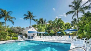 Piscina azul cristalina com espreguiçadeiras e guarda-sóis brancos, cercada por palmeiras e vegetação.