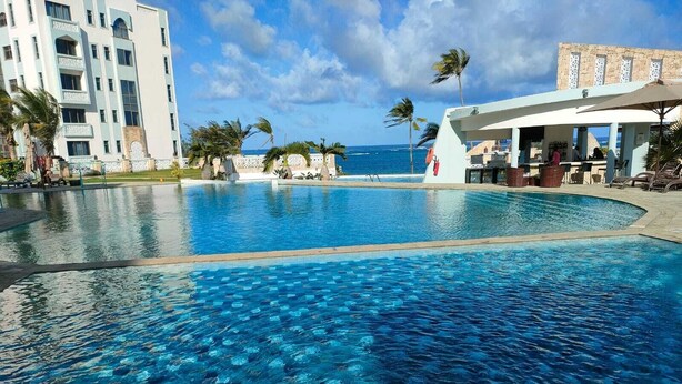 Uma piscina grande e convidativa, com borda curva, rodeada por espreguiçadeiras e um edifício com fachada azul clara.