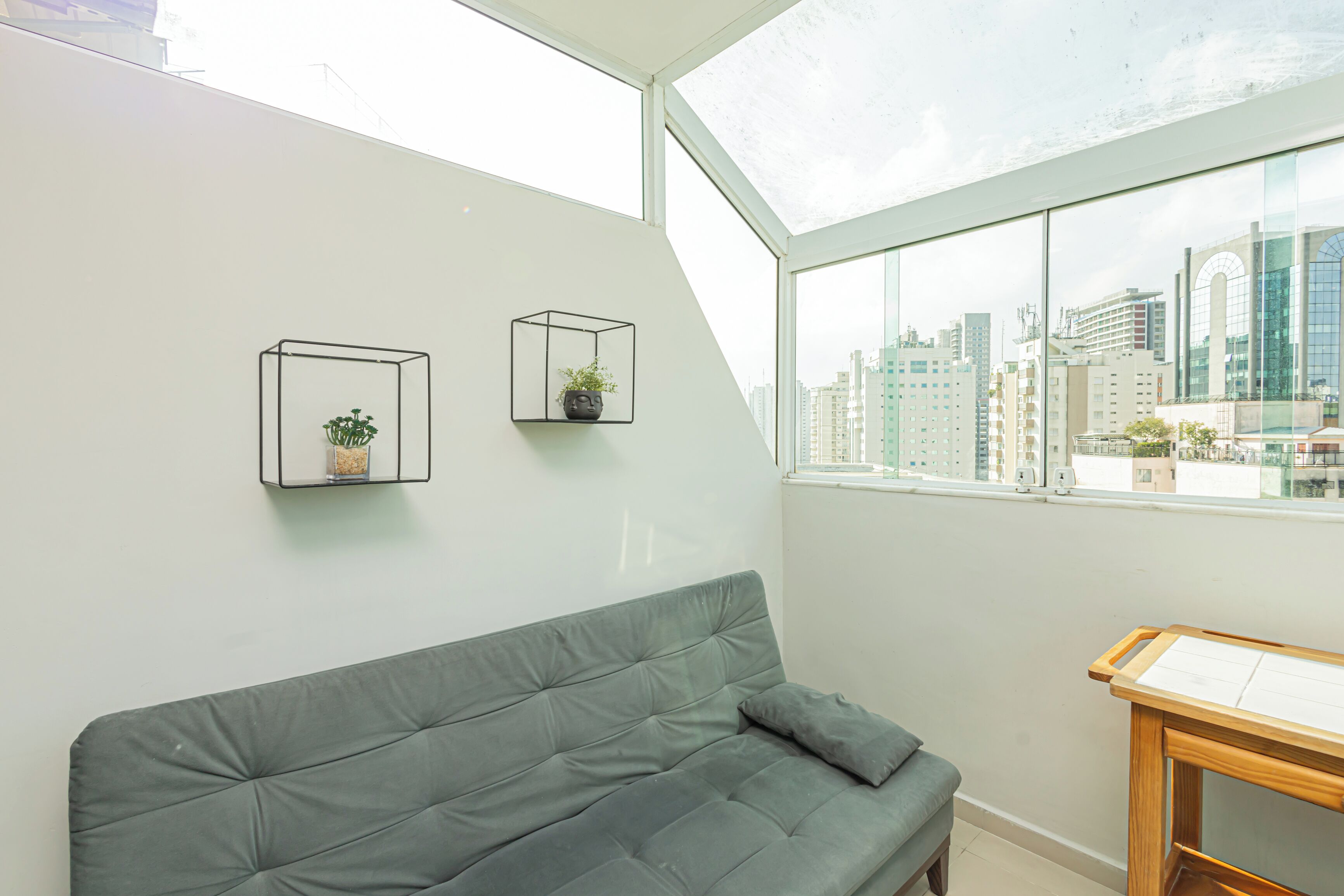 Uma sala de estar moderna com um sofá cinza, uma mesa lateral de madeira e duas prateleiras fixadas na parede com plantas. Uma grande janela oferece vista para a paisagem urbana.
