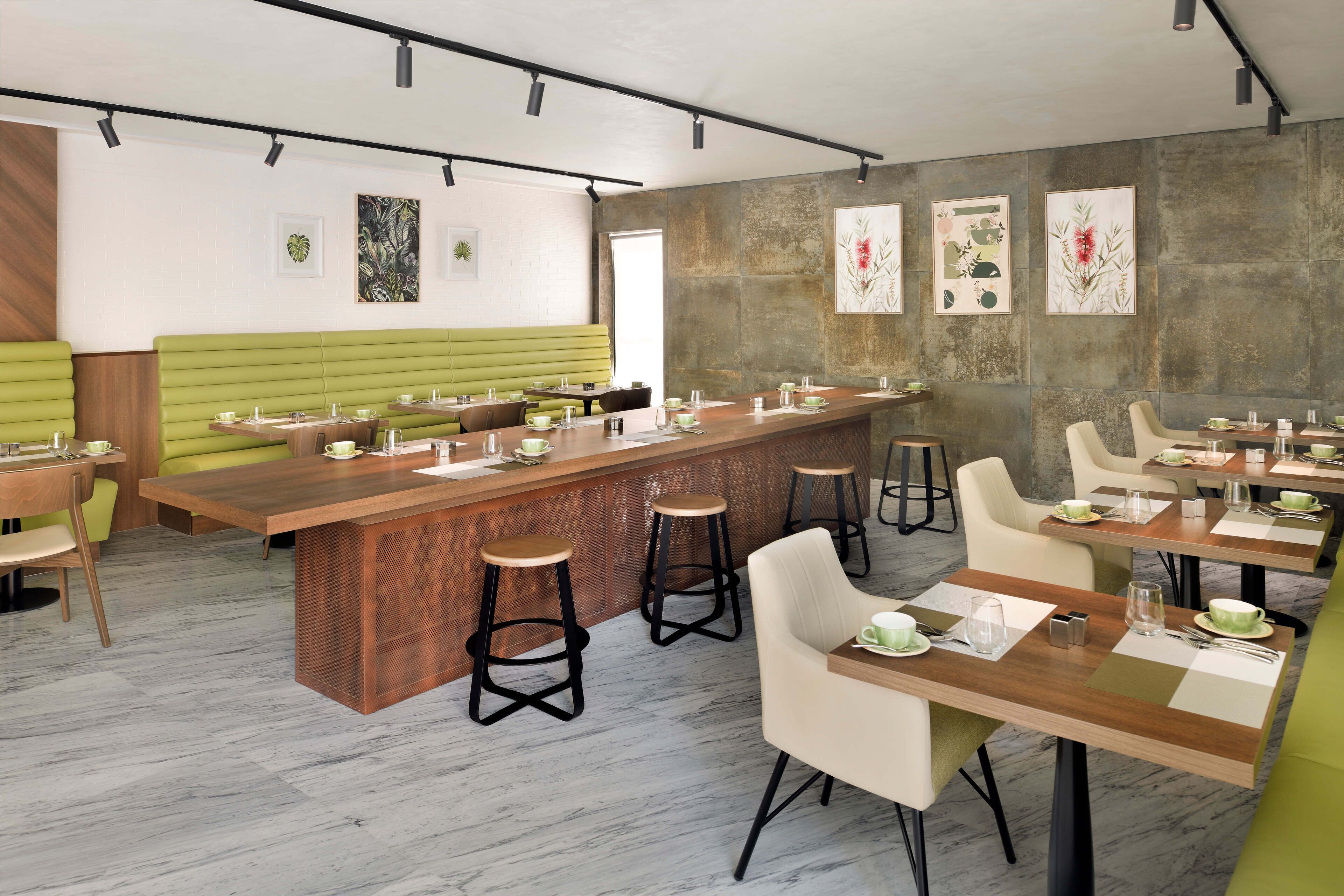 Um restaurante moderno com um balcão de madeira alongado, assentos estofados verdes e uma parede de pedra com impressões botânicas emolduradas.