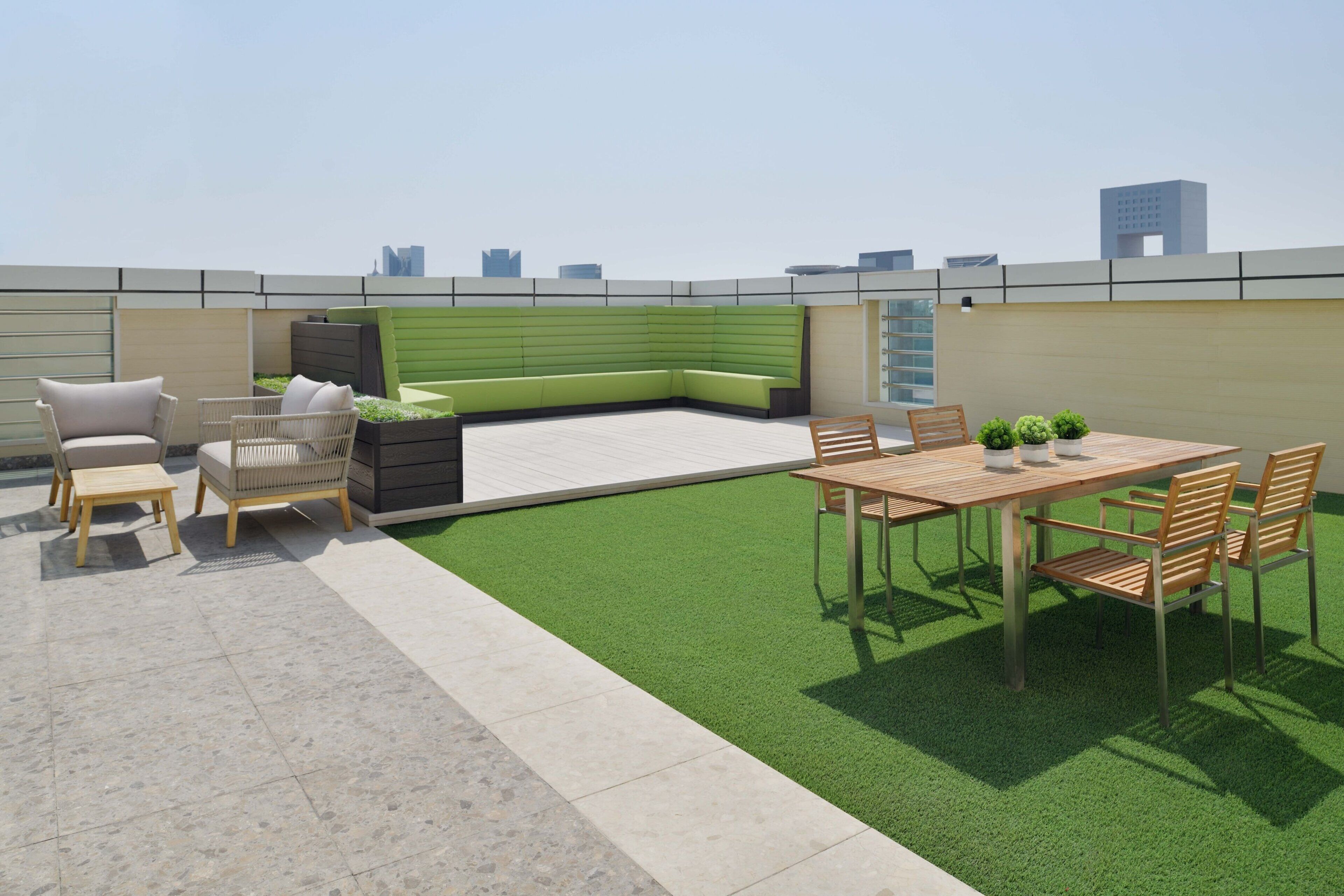 Um terraço no topo do prédio com grama sintética verde, uma mesa de madeira com cadeiras, um sofá e um espaço de estar com almofadas.