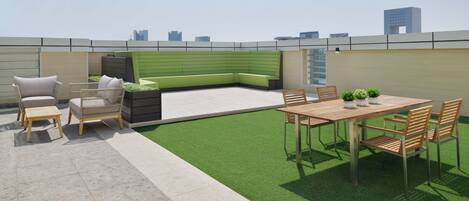 Um terraço no topo do prédio com grama sintética verde, uma mesa de madeira com cadeiras, um sofá e um espaço de estar com almofadas.