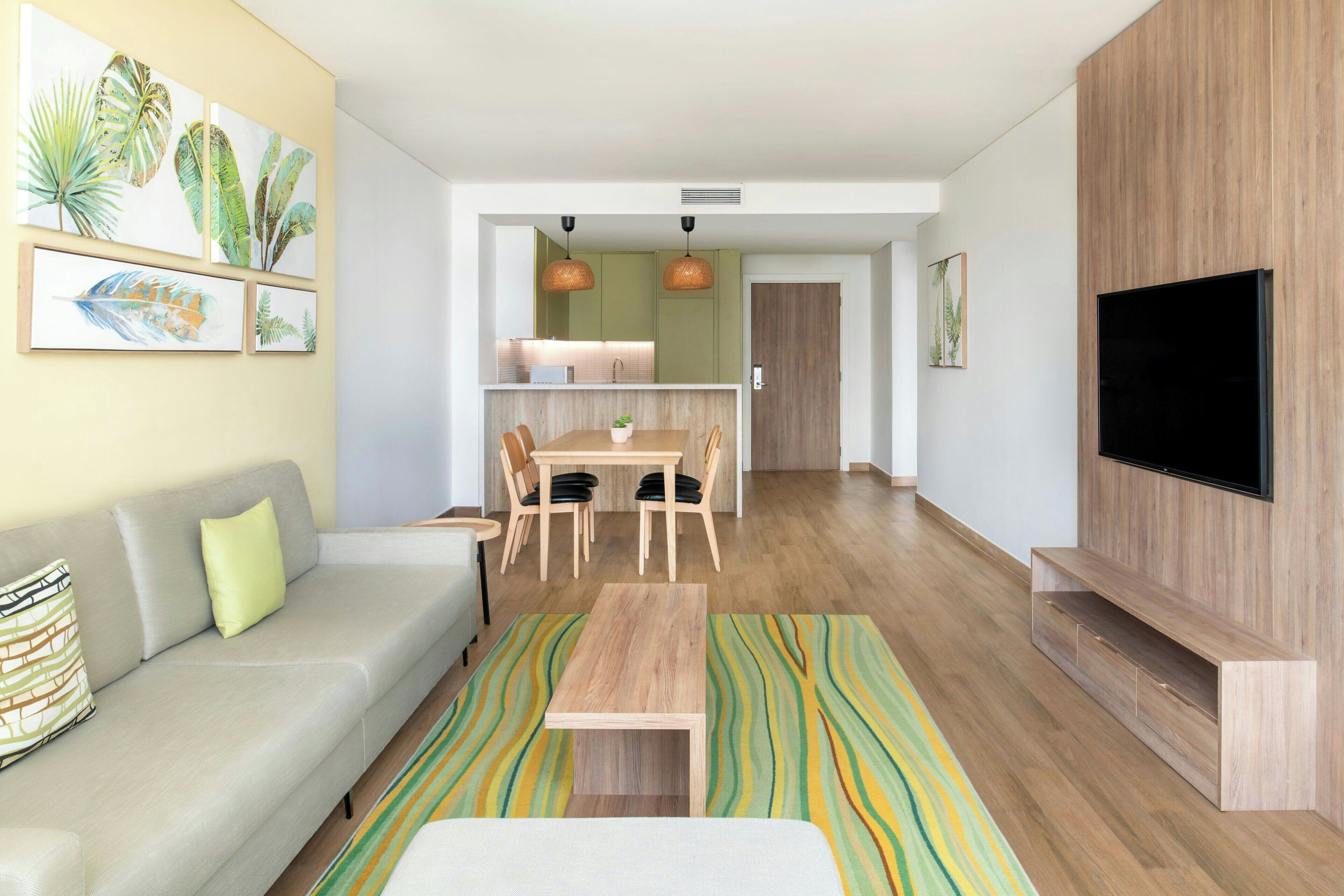 Uma sala de estar moderna com um sofá, uma mesa de jantar e cadeiras. Há uma televisão na parede e uma área de cozinha ao fundo.