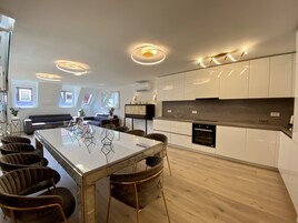 Cozinha moderna com armários brancos, forno preto e área de jantar com mesa de vidro e cadeiras escuras.
