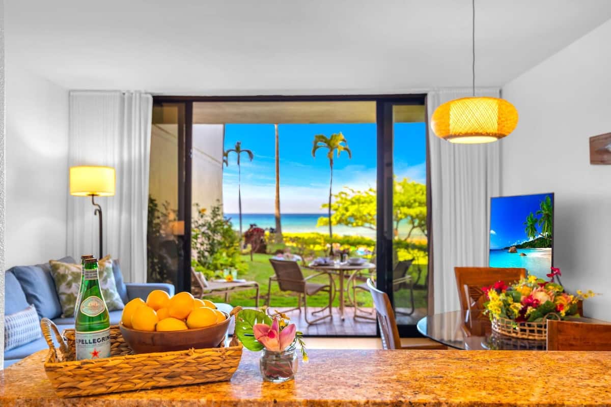 Quarto de hotel com vista para o oceano, área de refeições com mesa e cadeiras, e bar com uma garrafa e um recipiente de frutas.