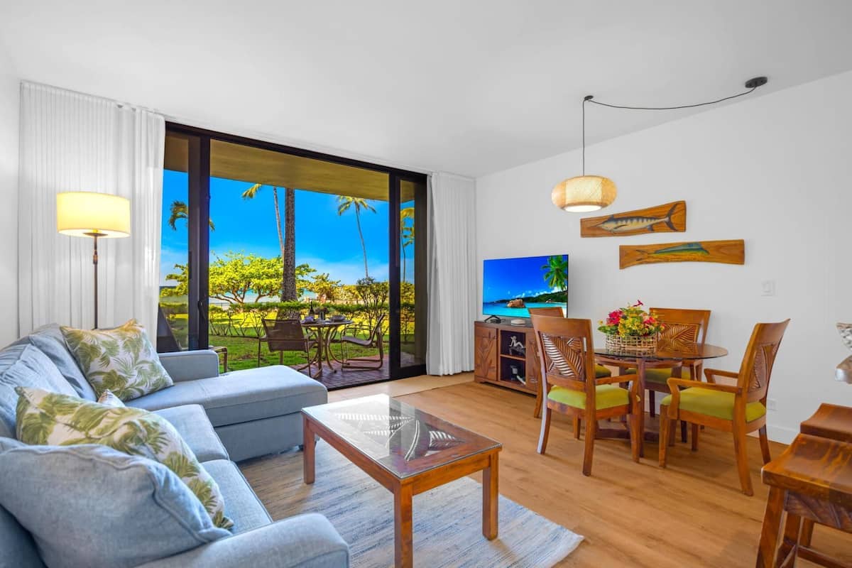 Uma sala de estar com sofá, mesa de centro, mesa de jantar com cadeiras, televisão e uma grande janela com vista.