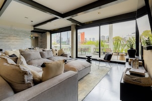 Uma sala de estar moderna com um amplo sofá em forma de L, uma área de jantar com mesa e cadeiras, e grandes janelas com vista para a cidade.