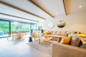 Uma sala de estar moderna com sofá, mesa de centro e área de jantar com cadeiras. Há janelas amplas que oferecem vista para o verde lá fora.