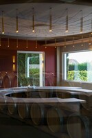 Um espaço com área de estar circular rodeada por barris de madeira, uma janela ampla com vista para o verde e luminárias pendentes.