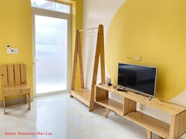 Um quarto claro com paredes amarelas, um banco de madeira, uma televisão sobre um móvel de madeira e uma porta que dá para o exterior.