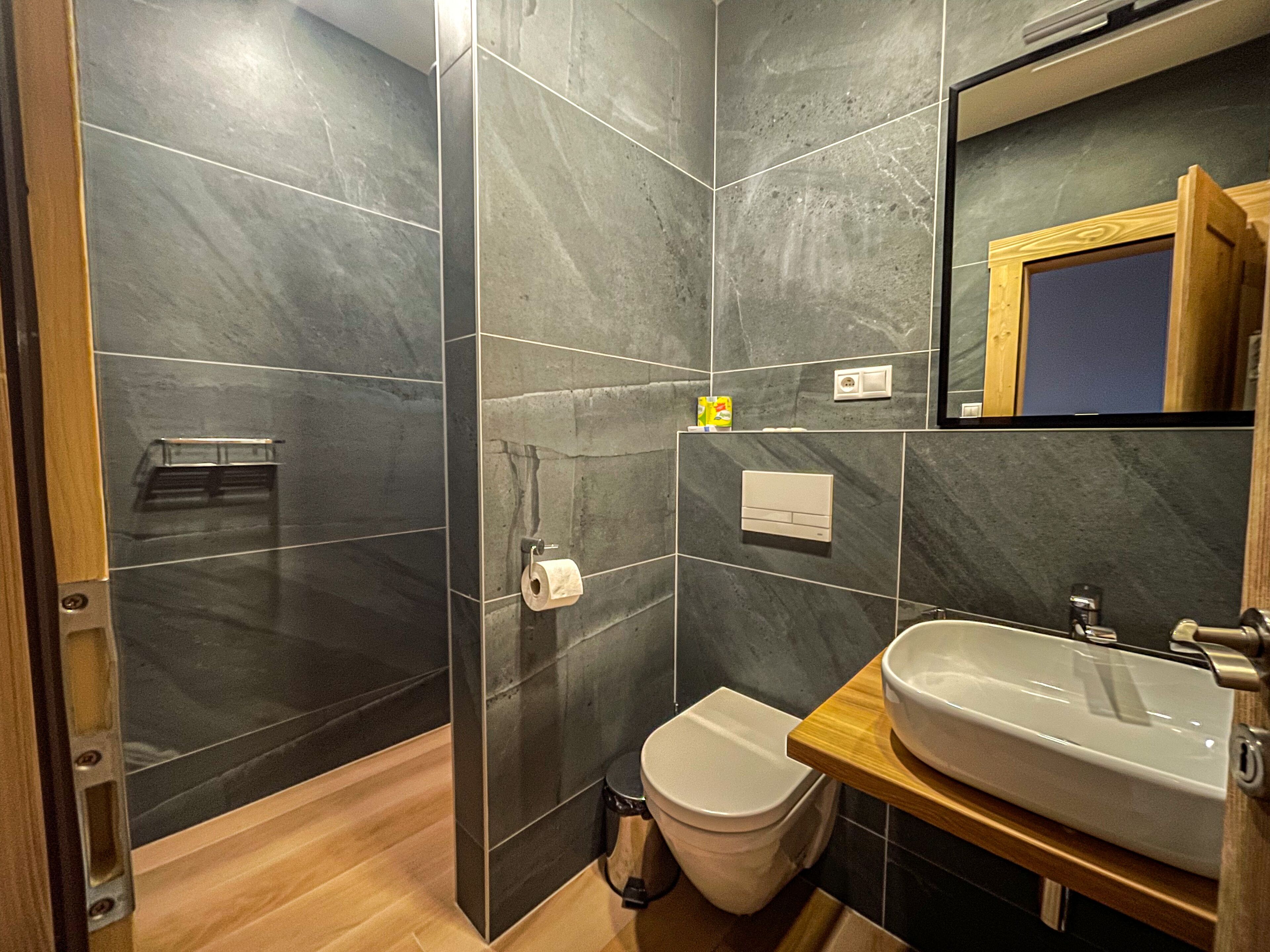 Uma casa de banho moderna com azulejos escuros, sanita, lavatório e espelho.