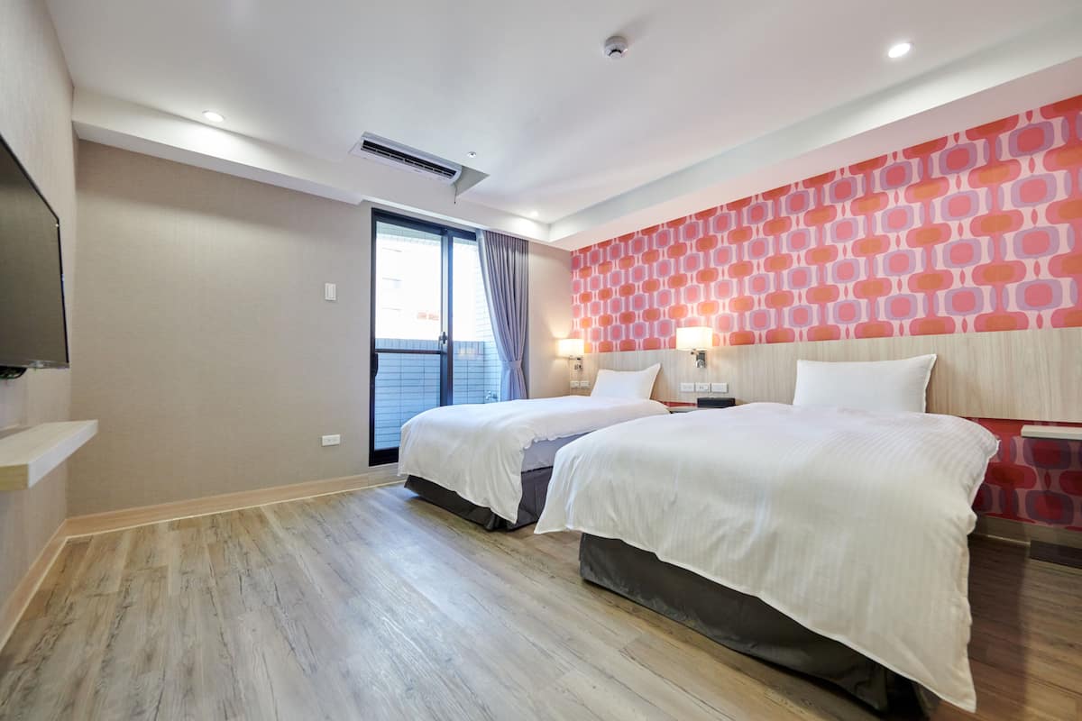 Quarto de hotel com duas camas, uma janela grande com cortinas, parede estampada e televisão de tela plana.
