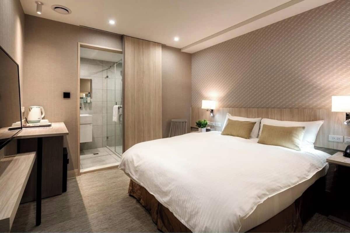 Quarto de hotel moderno com uma cama grande, mesas de cabeceira, uma escrivaninha, área de chuveiro e banheiro.
