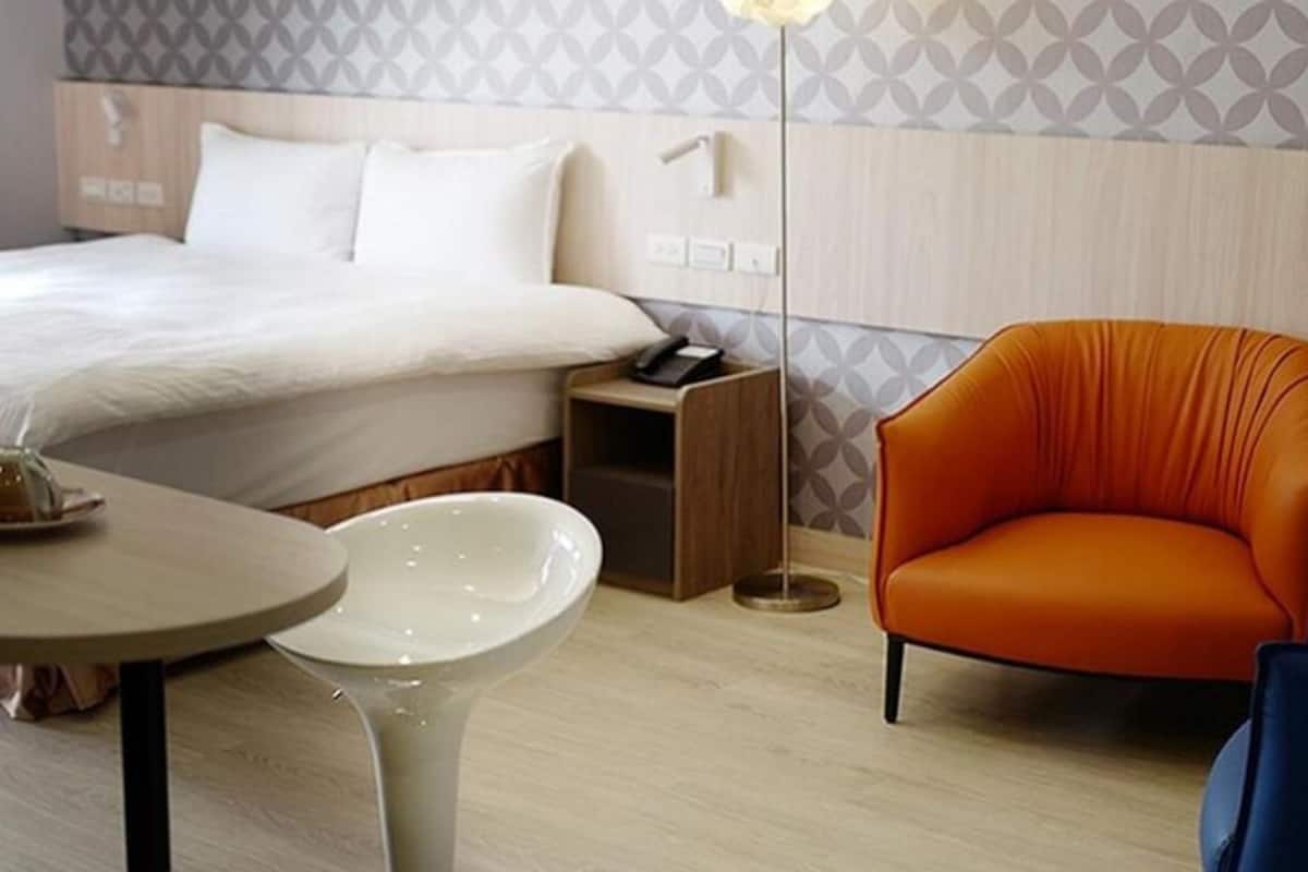 Quarto de hotel com cama, uma pequena mesa, uma cadeira branca, um poltrona laranja e uma cadeira azul.