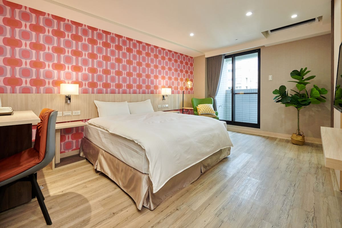 Quarto de hotel com cama, escrivaninha, cadeira, duas luminárias de parede, planta em vaso e uma janela grande.