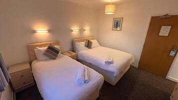 Quarto de hotel com duas camas, cada uma com cabeceira, mesas de cabeceira e luminárias. Há uma porta com fechadura e uma pequena janela.