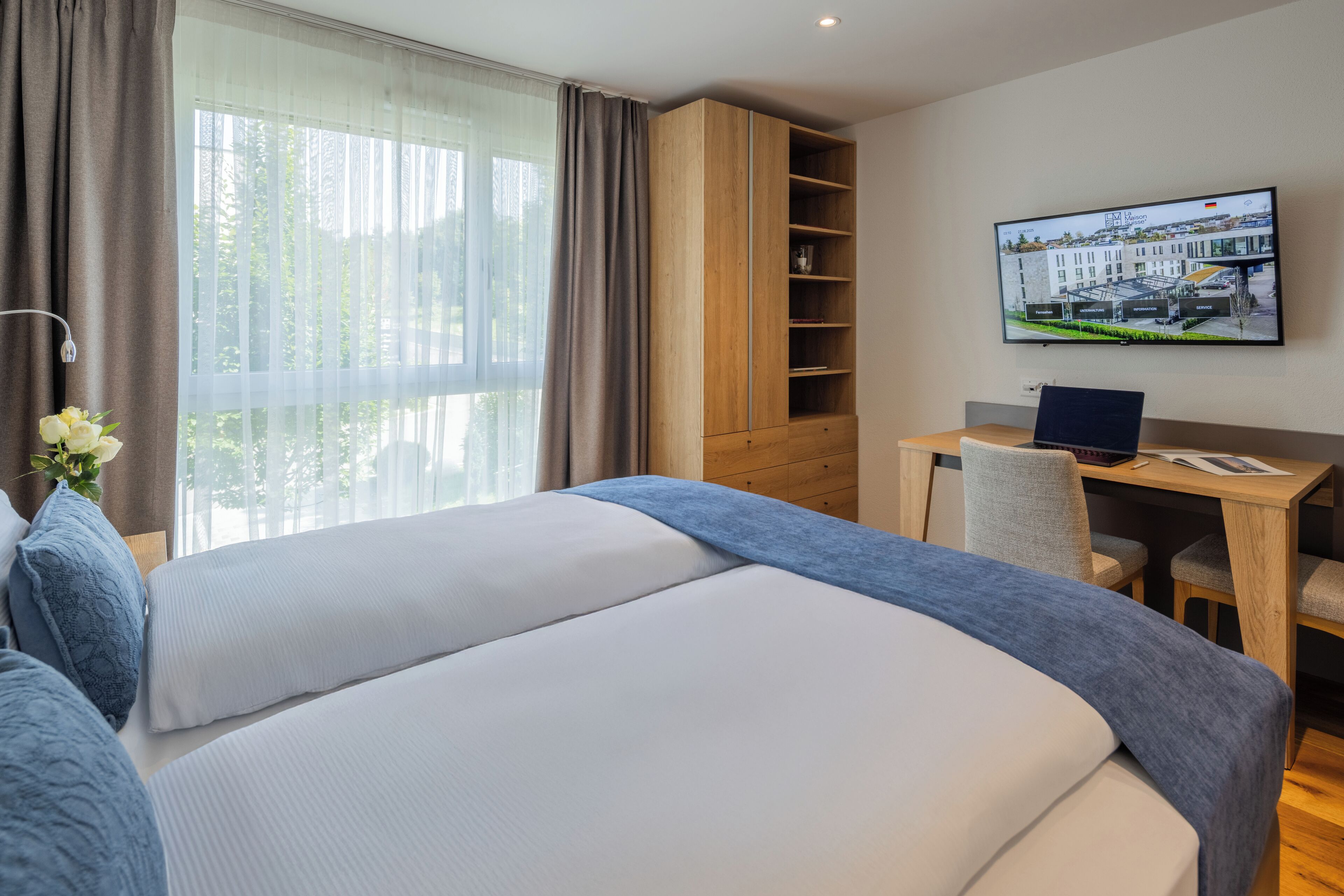 Quarto de hotel com uma cama grande, uma escrivaninha com um laptop, uma televisão na parede e uma janela com cortinas.