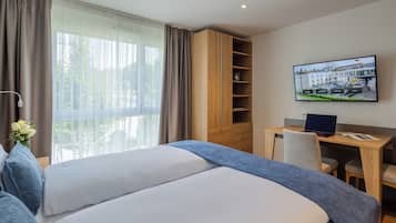 Quarto de hotel com uma cama grande, uma escrivaninha com um laptop, uma televisão na parede e uma janela com cortinas.