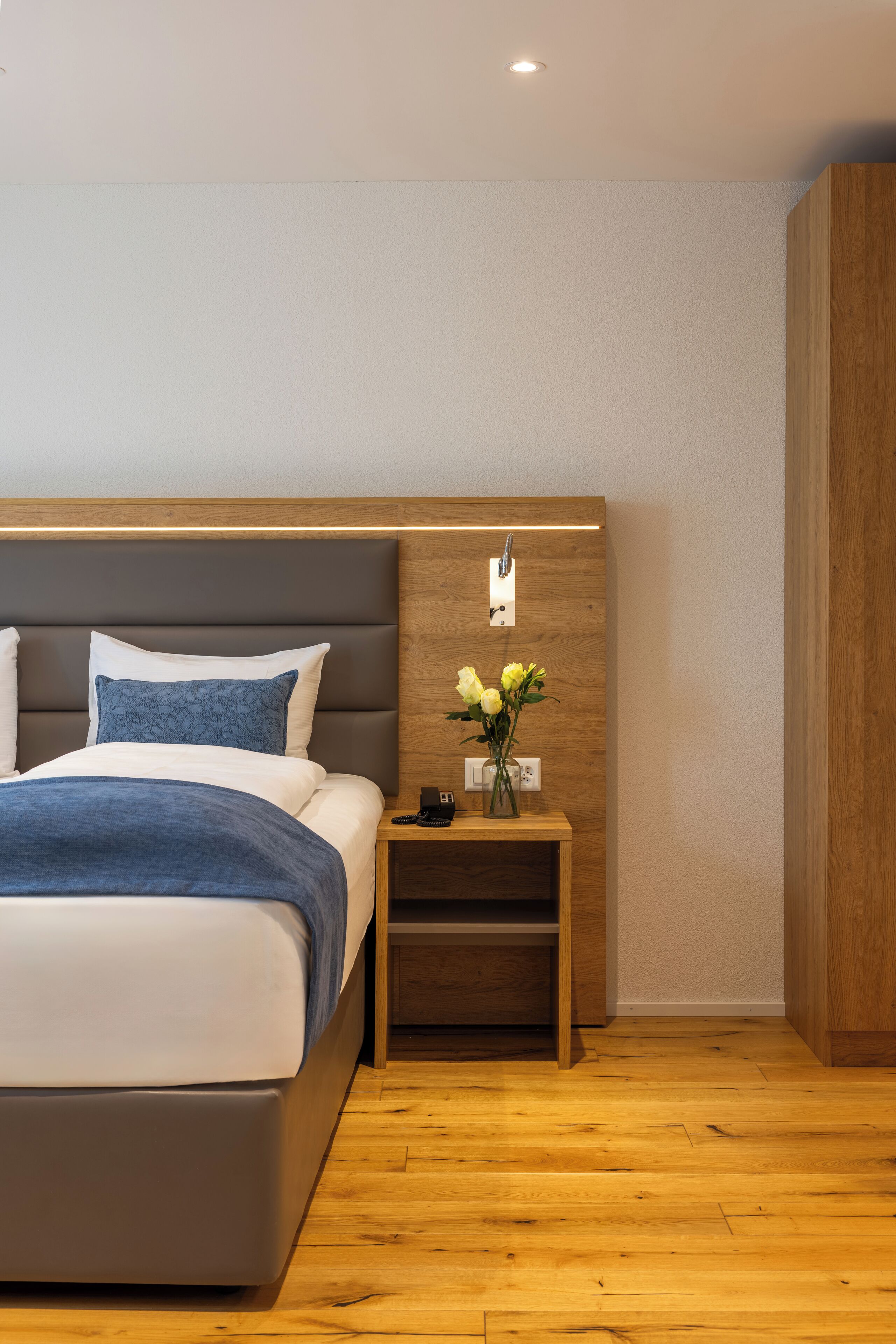 Quarto de hotel moderno com uma cama grande, uma mesa de cabeceira de madeira com um vaso de flores e um guarda-roupa de madeira.