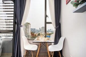 Uma área de refeições com uma mesa posta para dois, duas cadeiras brancas e vista para a cidade através de uma janela com cortinas.