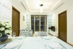 Uma área de refeições com uma mesa branca posta para quatro pessoas, um sofá e uma televisão ao fundo.