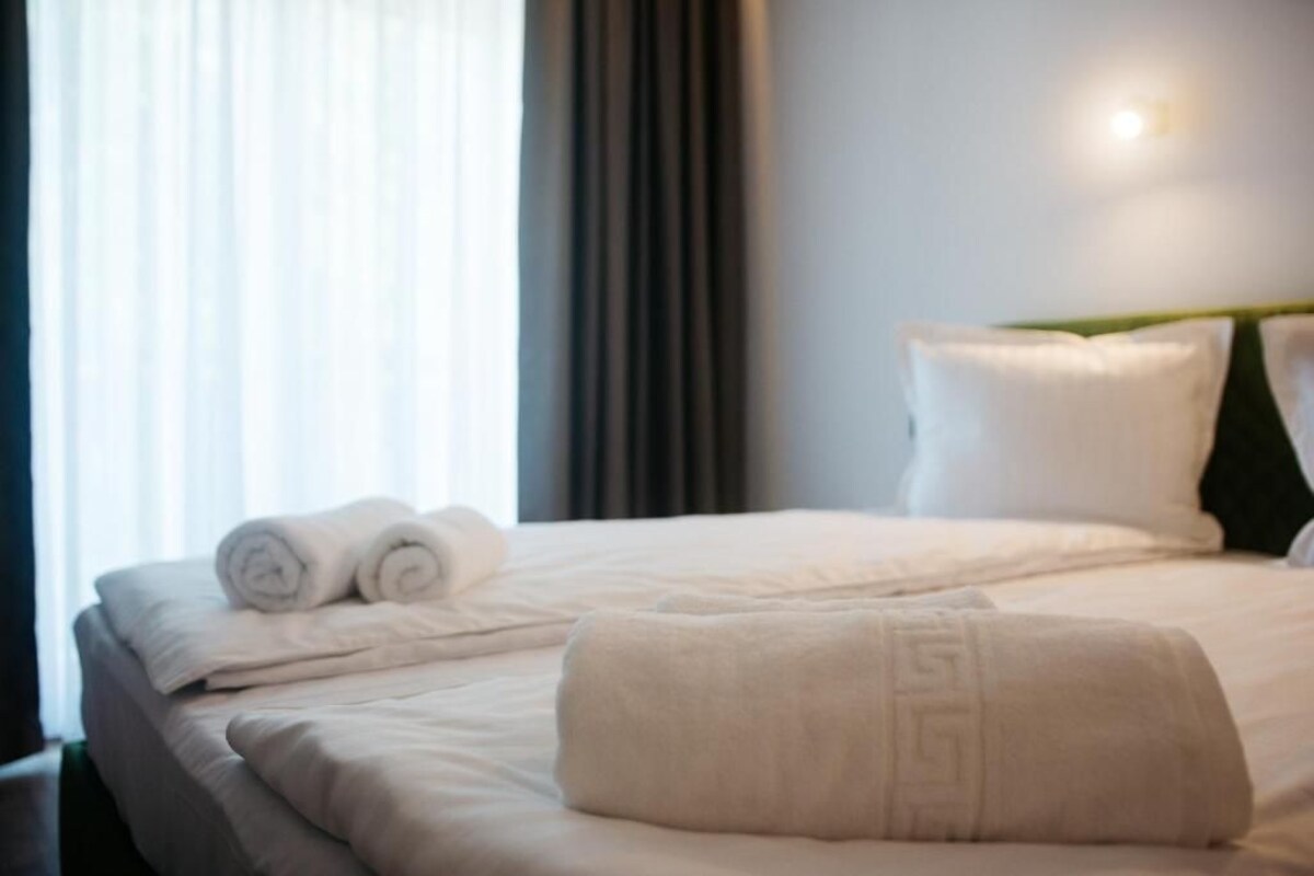 Cama bem arrumada com toalhas brancas dobradas, cabeceira e luminária fixada na parede.