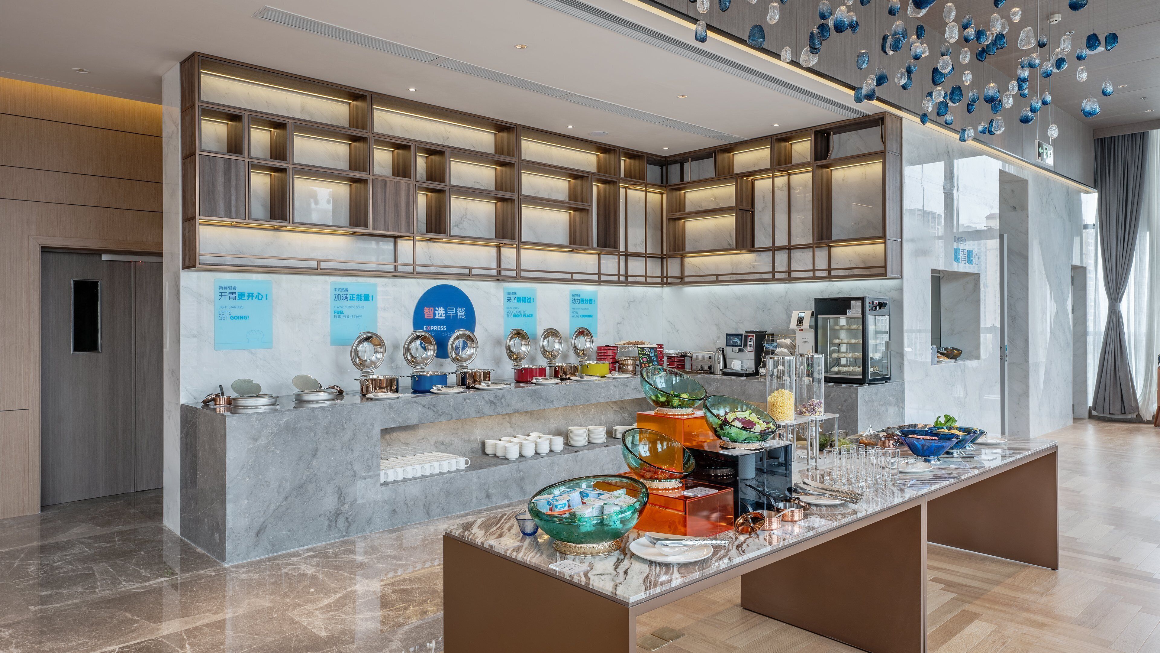 Espaço de pequeno-almoço de um hotel moderno, com bancadas de mármore, diversas opções de refeições e uma máquina de café digital.