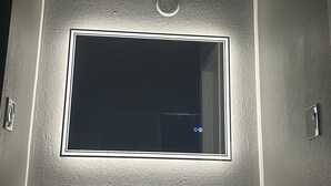 Uma casa de banho com um espelho retangular sobre uma pia, um candeeiro na parede e uma maçaneta.