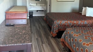 Quarto de hotel com piso de madeira, cama com colcha estampada, mesa de cabeceira, espelho, quadro na parede e televisão fixada na parede.