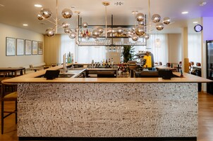 Uma área de bar com ilha central, banquetas, caixa registradora e diversos frascos e copos.