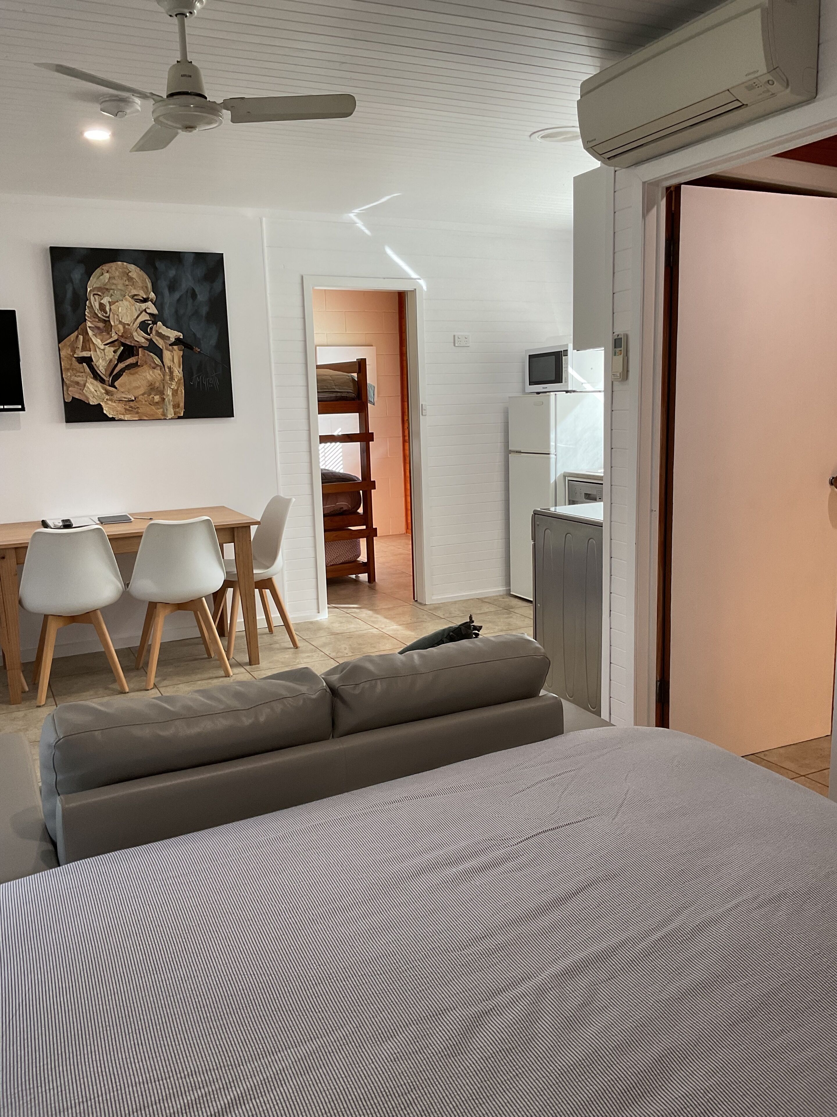 Quarto com cama, escrivaninha e cadeira. Há um quadro na parede e um ventilador de teto.