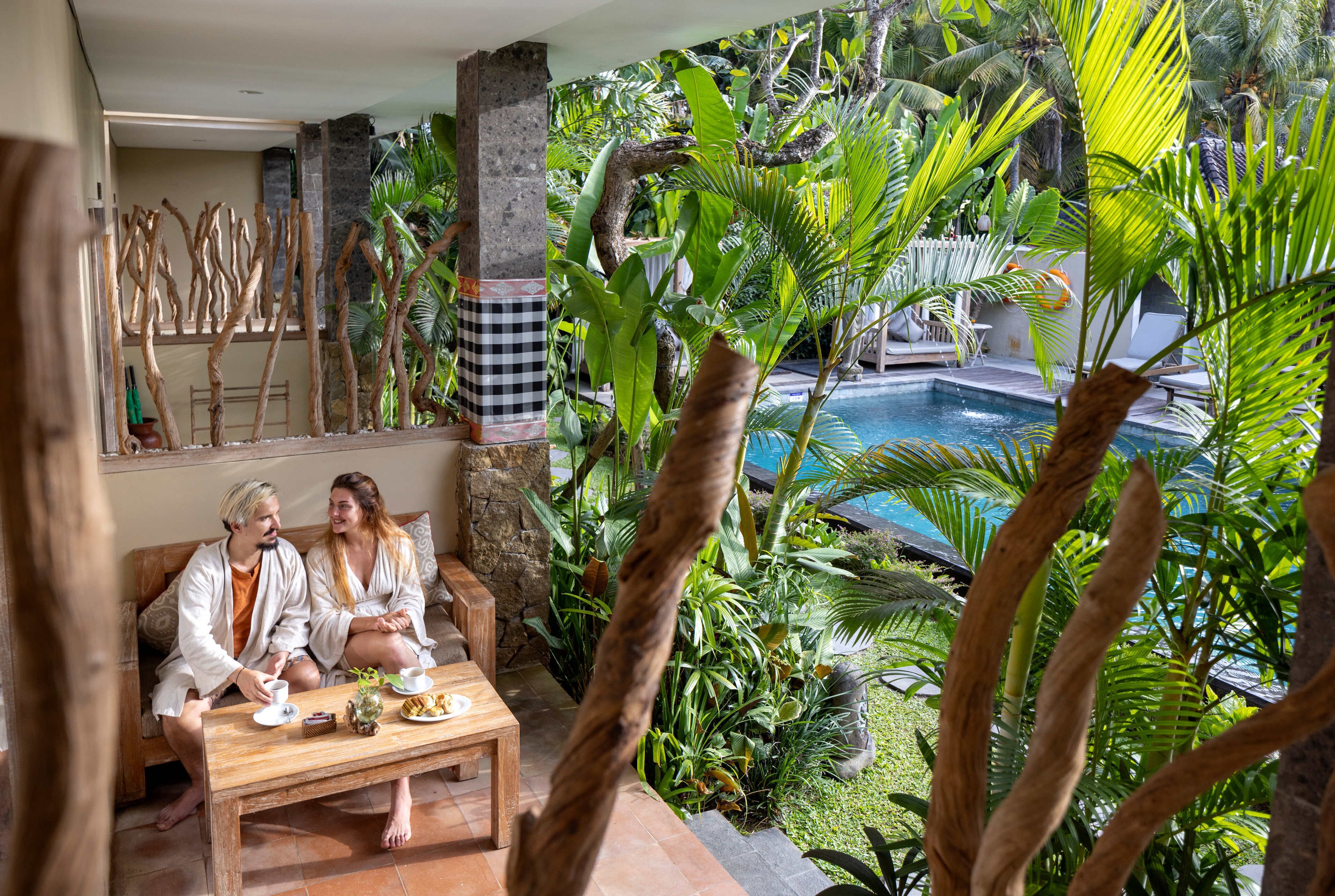 Um casal sentado em um banco de madeira em um cenário tropical, com piscina e vegetação exuberante.