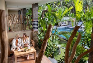 Um casal sentado em um banco de madeira em um cenário tropical, com piscina e vegetação exuberante.