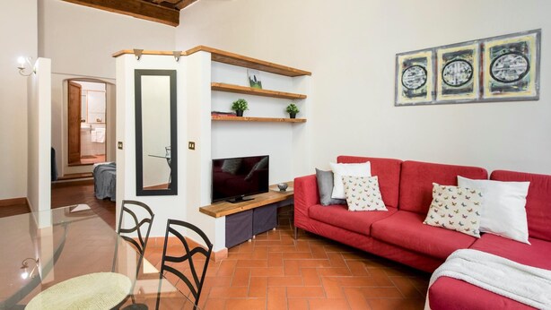 Uma sala de estar com um sofá vermelho, uma televisão, um móvel de madeira para TV, um espelho e um espaço de jantar com uma mesa e cadeiras de vidro.