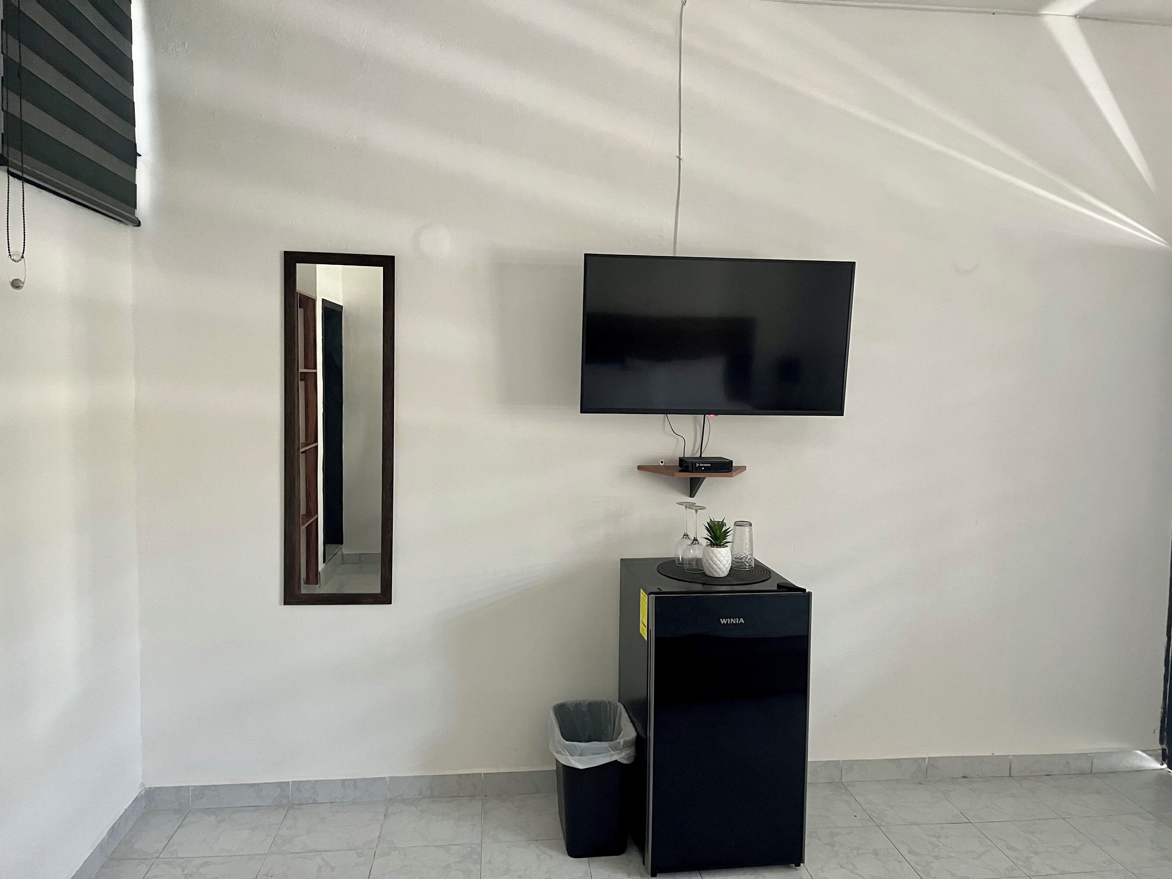 Uma sala de estar moderna com uma TV de tela plana na parede, uma pequena prateleira de madeira com um alto-falante, um rack de entretenimento preto com uma TV e uma lixeira.