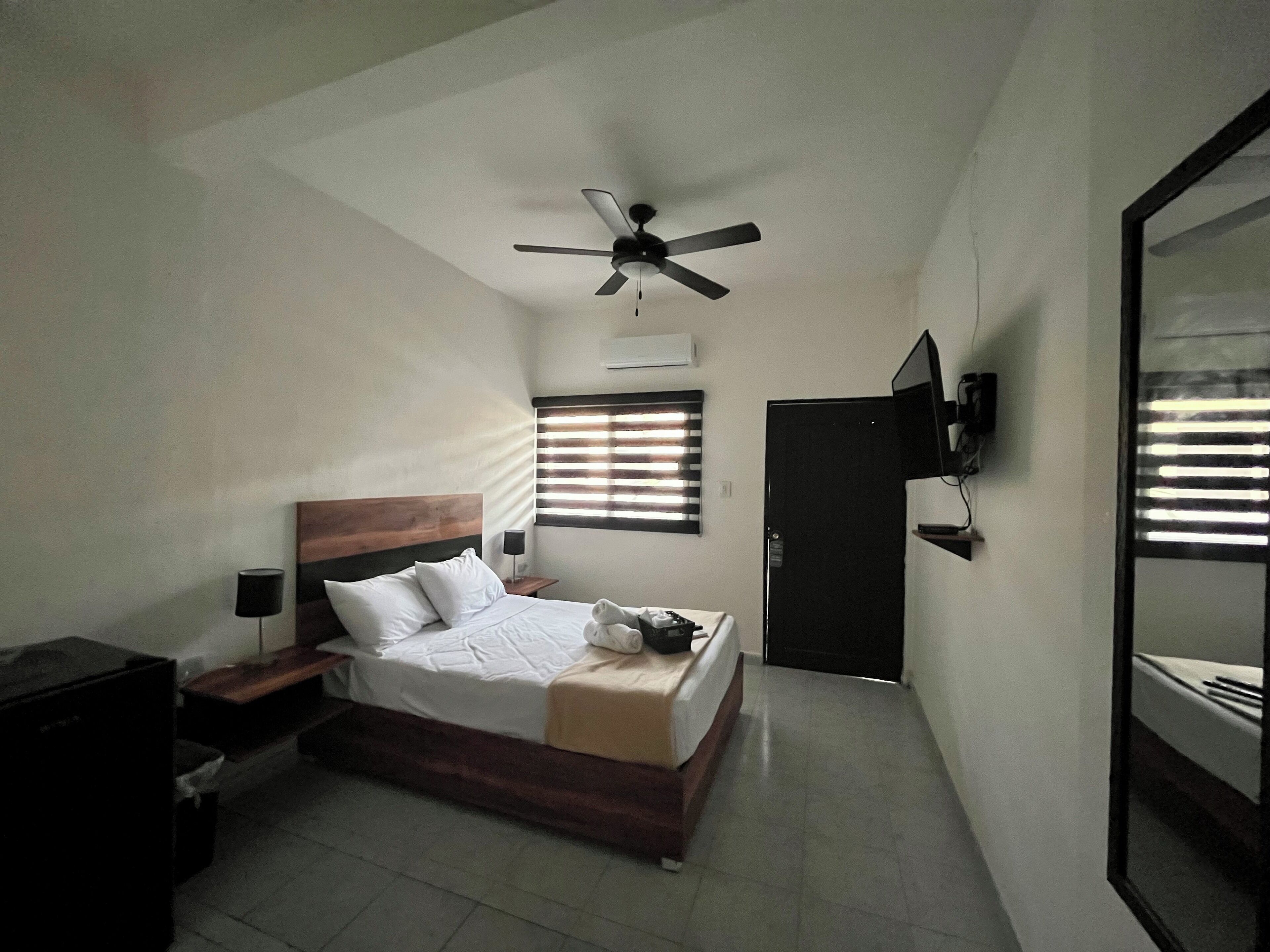 Quarto com cama, mesa de cabeceira, escrivaninha, ventoinha no teto, janela com persianas, porta e TV fixada na parede.