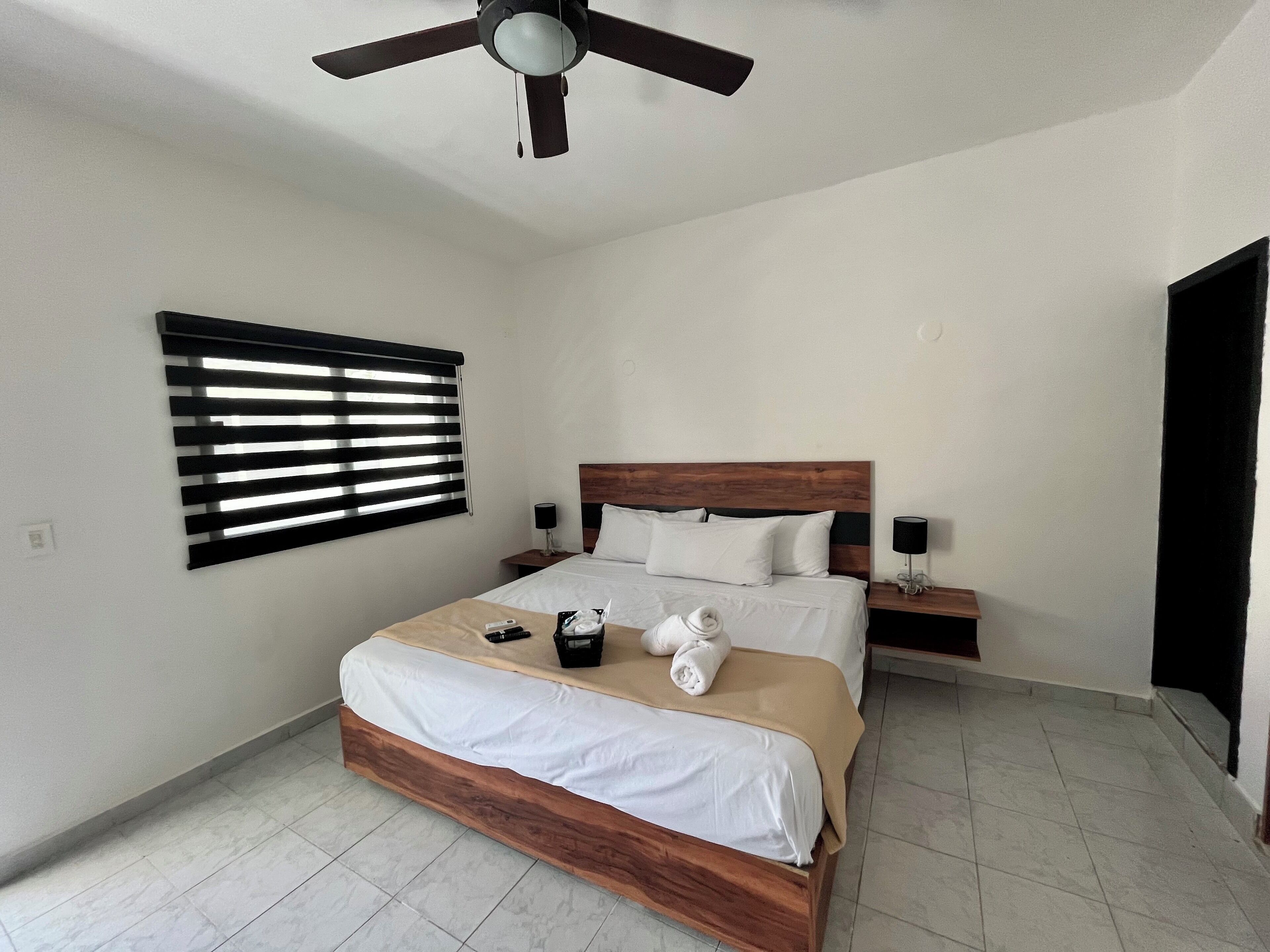 Um quarto com cama, ventilador de teto, janela com persianas e mesa de cabeceira com abajur.