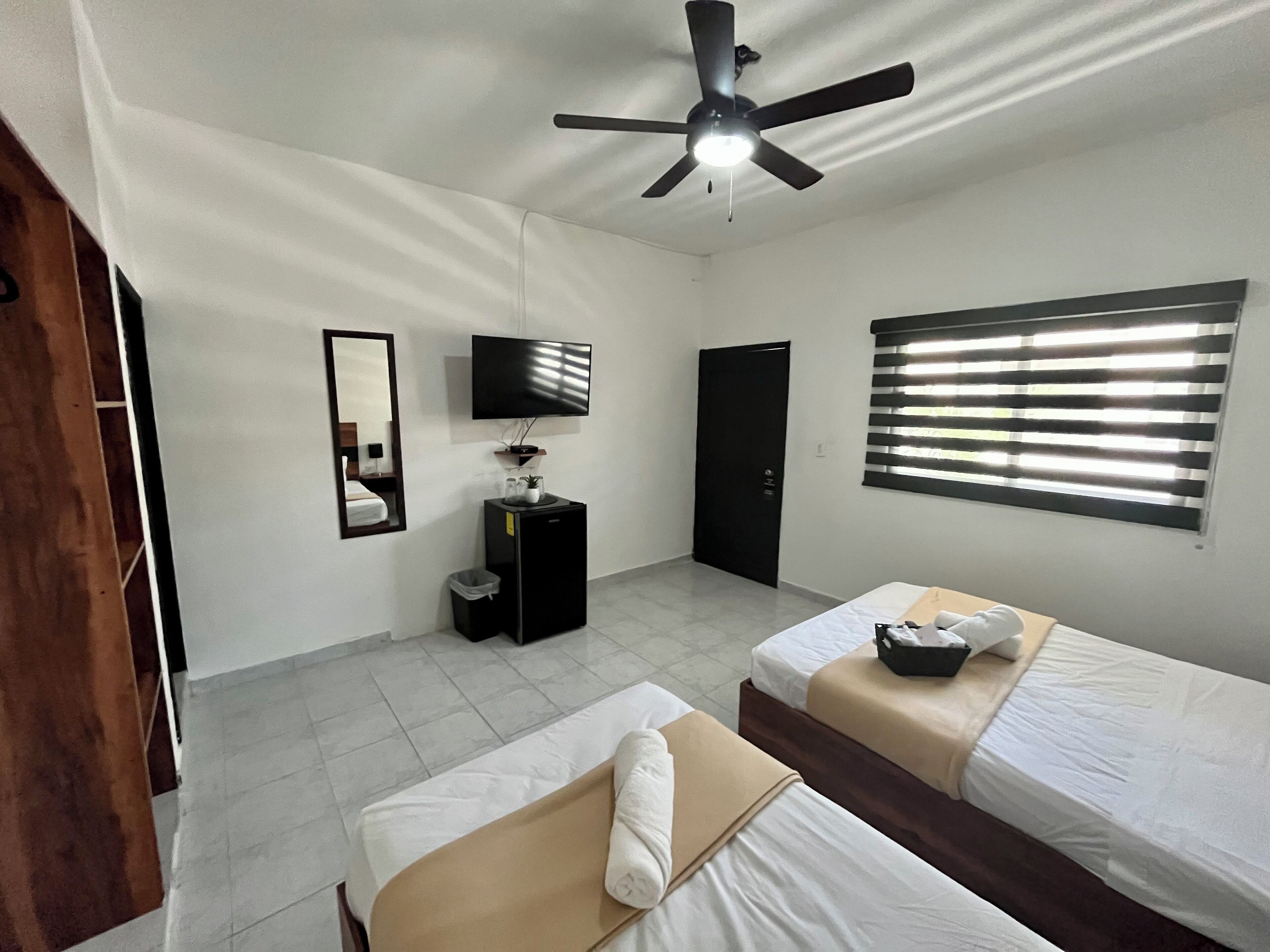 Quarto de hotel com duas camas, televisão, ventoinha no teto, porta e janela com persianas.