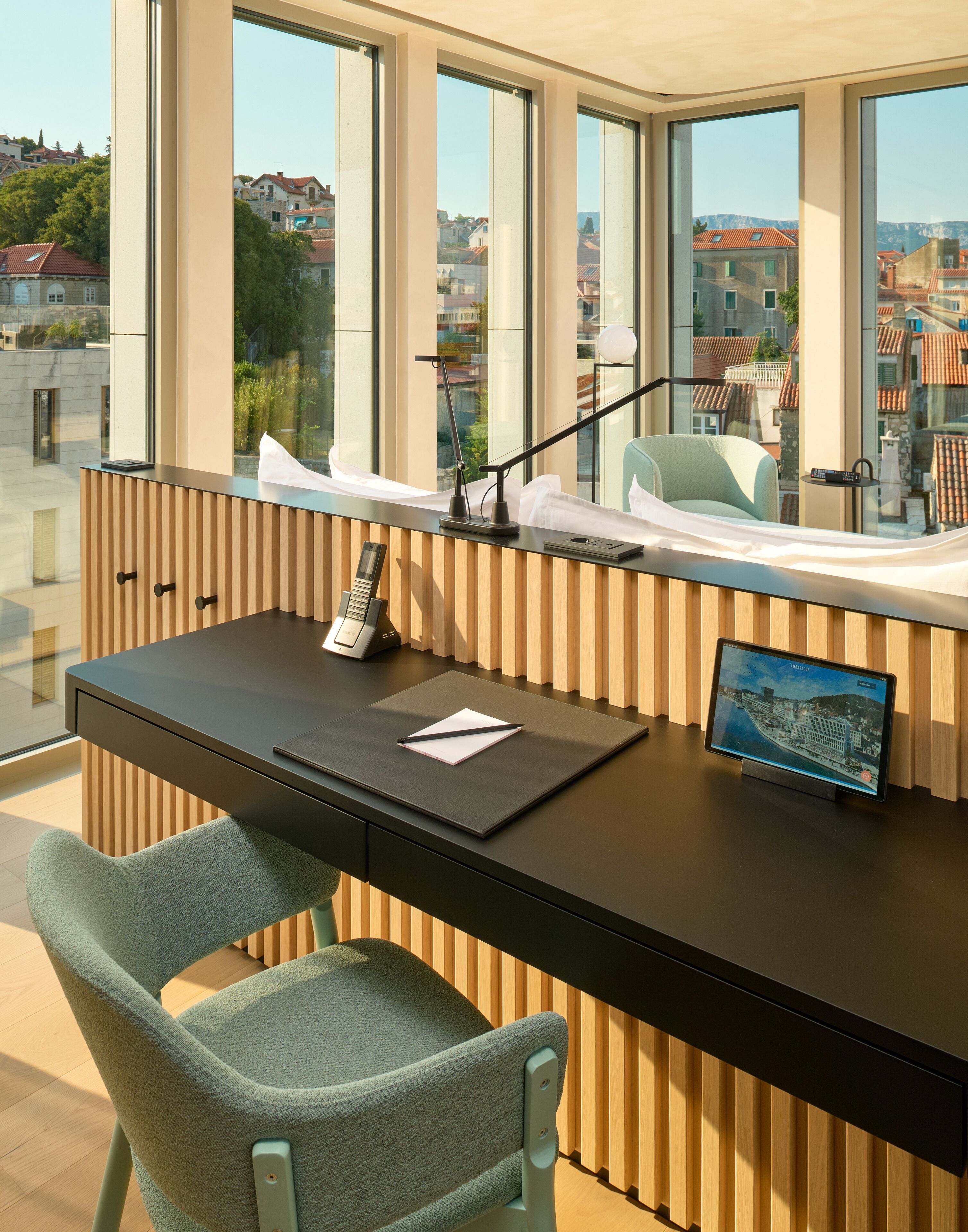 Uma escrivaninha moderna com um laptop, um smartphone e um bloco de notas. Há uma cadeira em frente à escrivaninha e uma janela com vista para edifícios e árvores.